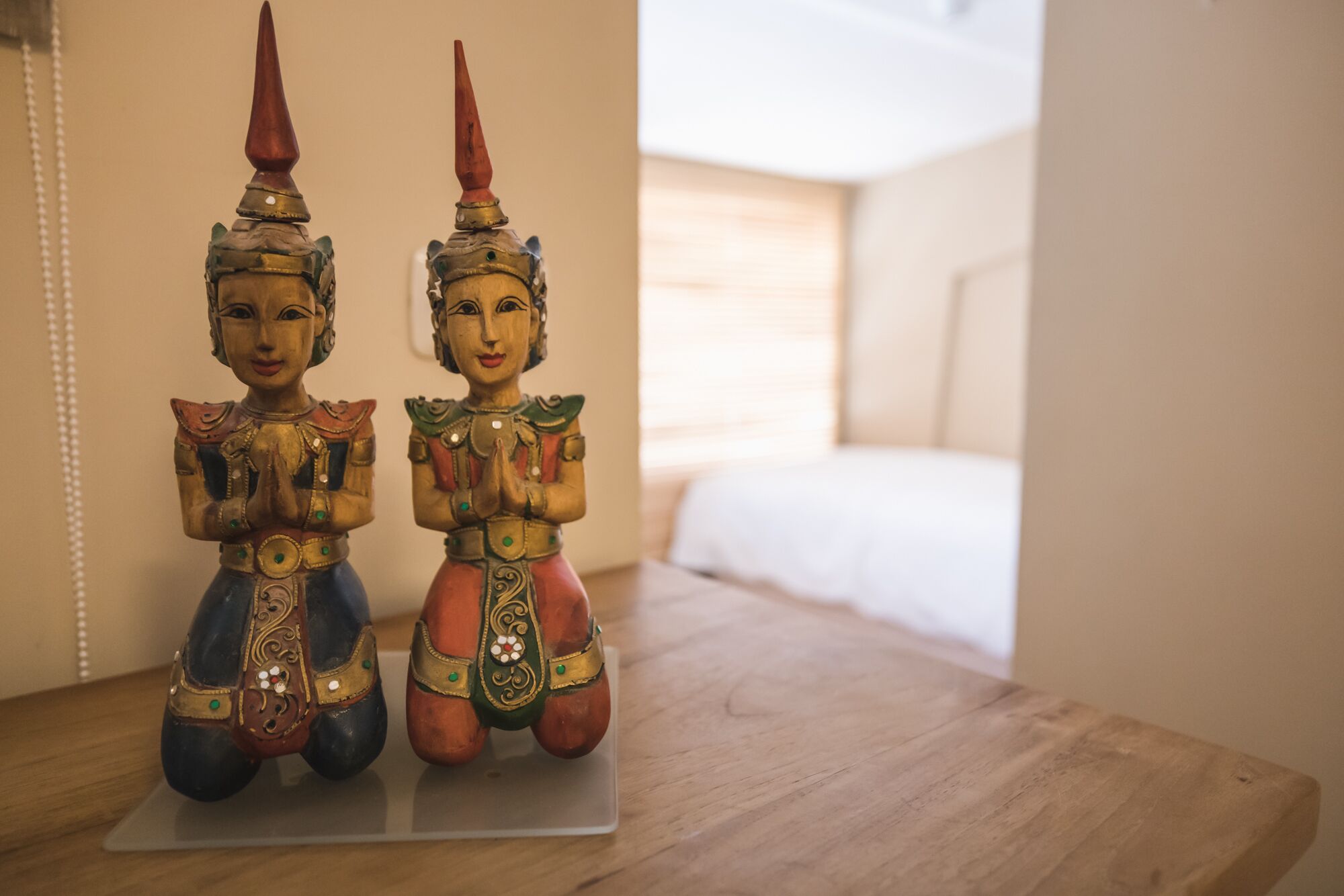 Duas figuras de madeira ornamentadas sobre um suporte de vidro transparente em um quarto com uma cama ao fundo.