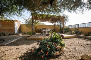 Área de varanda coberta com pérgola de madeira, mesa e cadeiras para refeições. Há plantas em vasos e um pequeno jardim com flores.