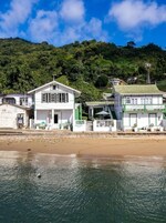 Um edifício costeiro com fachada branca e detalhes verdes, localizado à beira de uma praia arenosa com um cenário de vegetação exuberante.