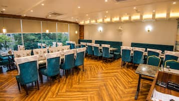 Restaurante com piso de madeira em padrão herringbone, cadeiras estofadas em tom de azul-esverdeado e mesas postas com toalhas brancas.