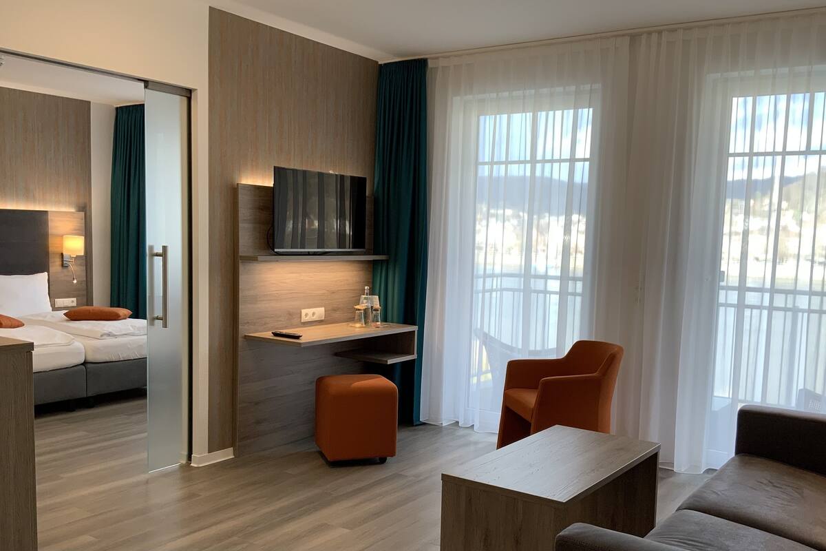 Quarto de hotel moderno com cama, escrivaninha, TV e área de estar com duas cadeiras.