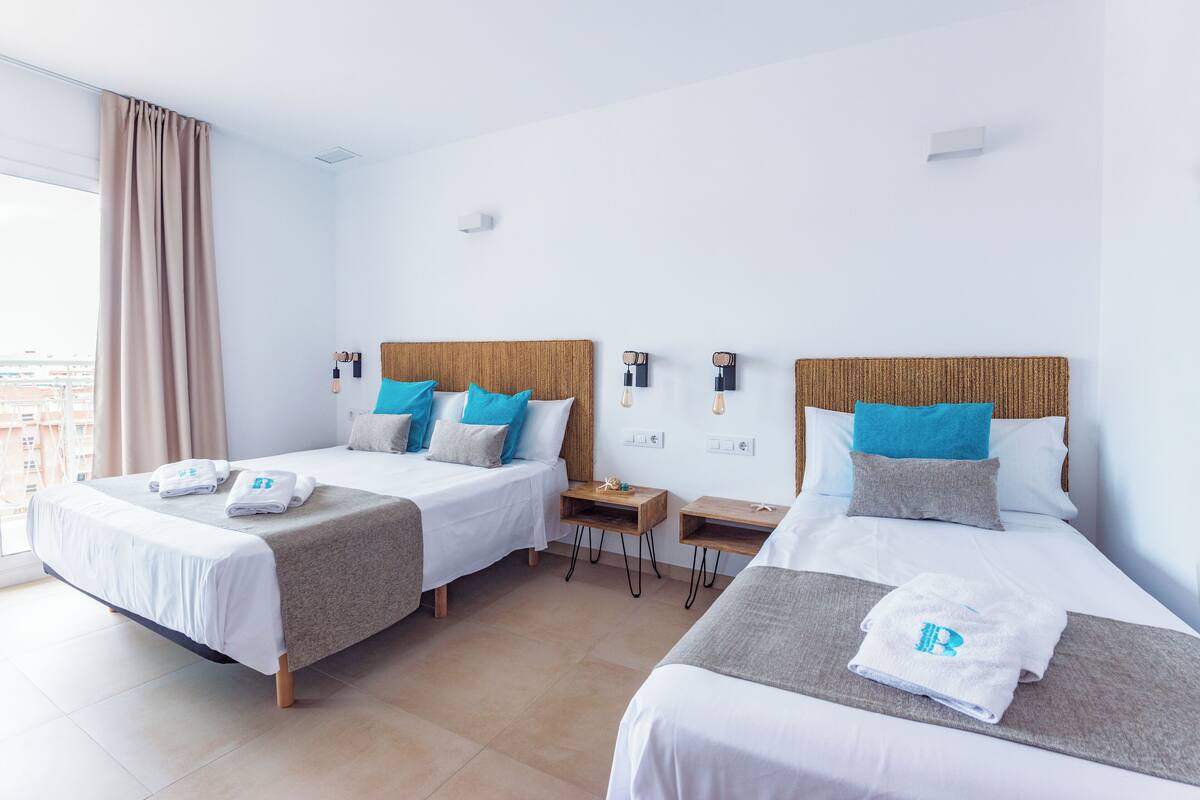 Quarto de hotel com duas camas, cada uma com colcha branca, manta cinza e almofadas azuis e cinzas. Há duas mesas de cabeceira com luminárias, uma janela com cortinas e vista para edifícios.