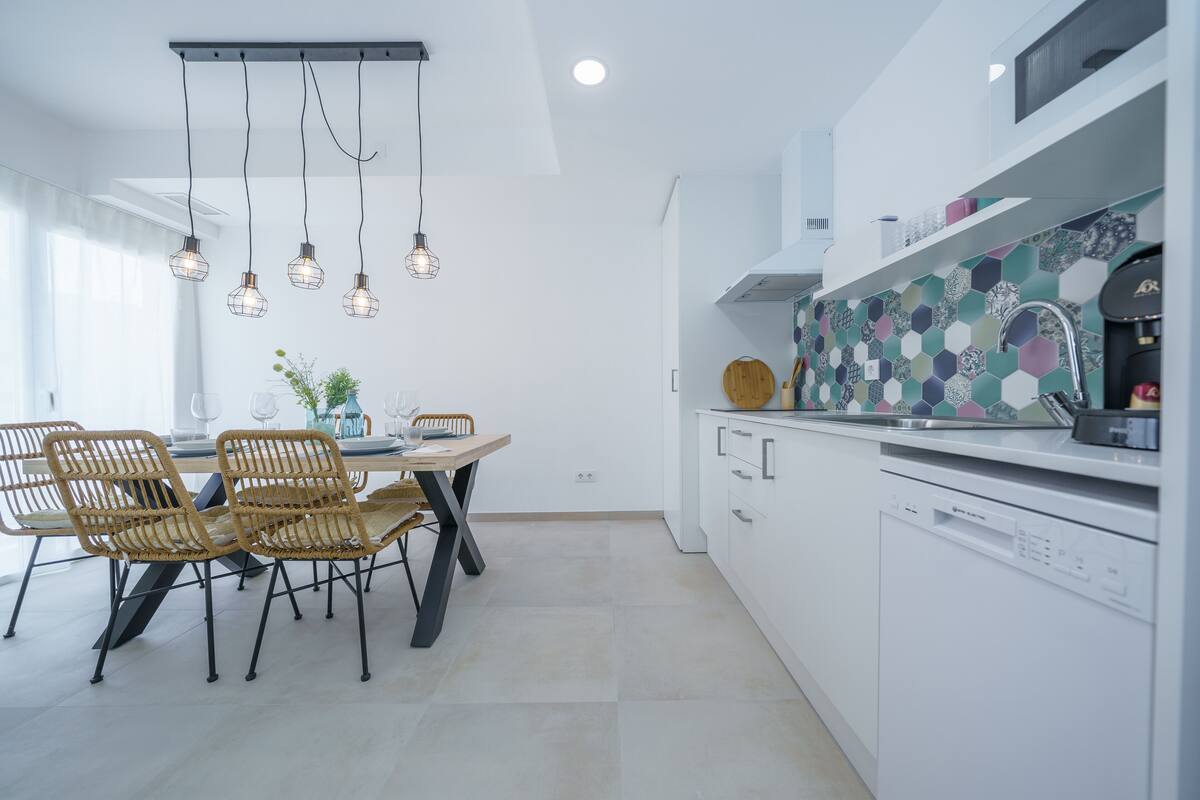 Cozinha moderna com armários brancos, área de jantar com cadeiras de vime e uma moldura de azulejos coloridos em formato hexagonal.