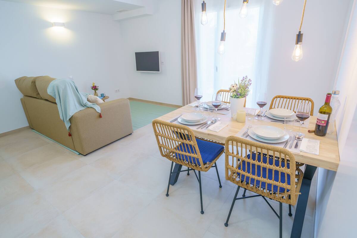 Uma sala de jantar com mesa de madeira posta para quatro pessoas, cercada por cadeiras de vime com almofadas azuis.