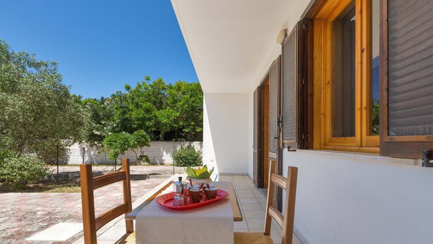 Um pátio com uma mesa posta para dois, cadeiras de madeira e um tabuleiro vermelho com xícaras e uma colher.