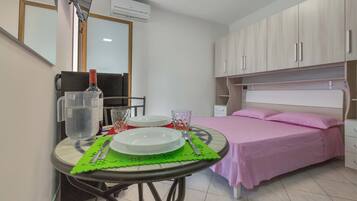 Quarto com cama, mesa de cabeceira com colcha rosa, espelho e janela.