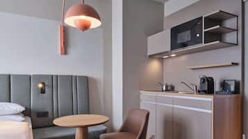 Quarto de hotel moderno com um sofá cinzento, uma mesa redonda pequena, uma poltrona marrom, uma cozinha com micro-ondas e uma luminária pendente.