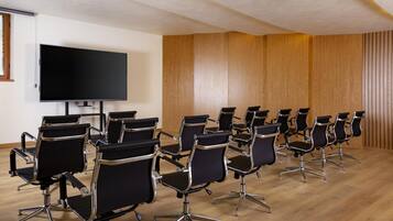 Uma sala de conferências com fileiras de cadeiras de escritório pretas, uma televisão de tela plana grande e painéis de madeira nas paredes.