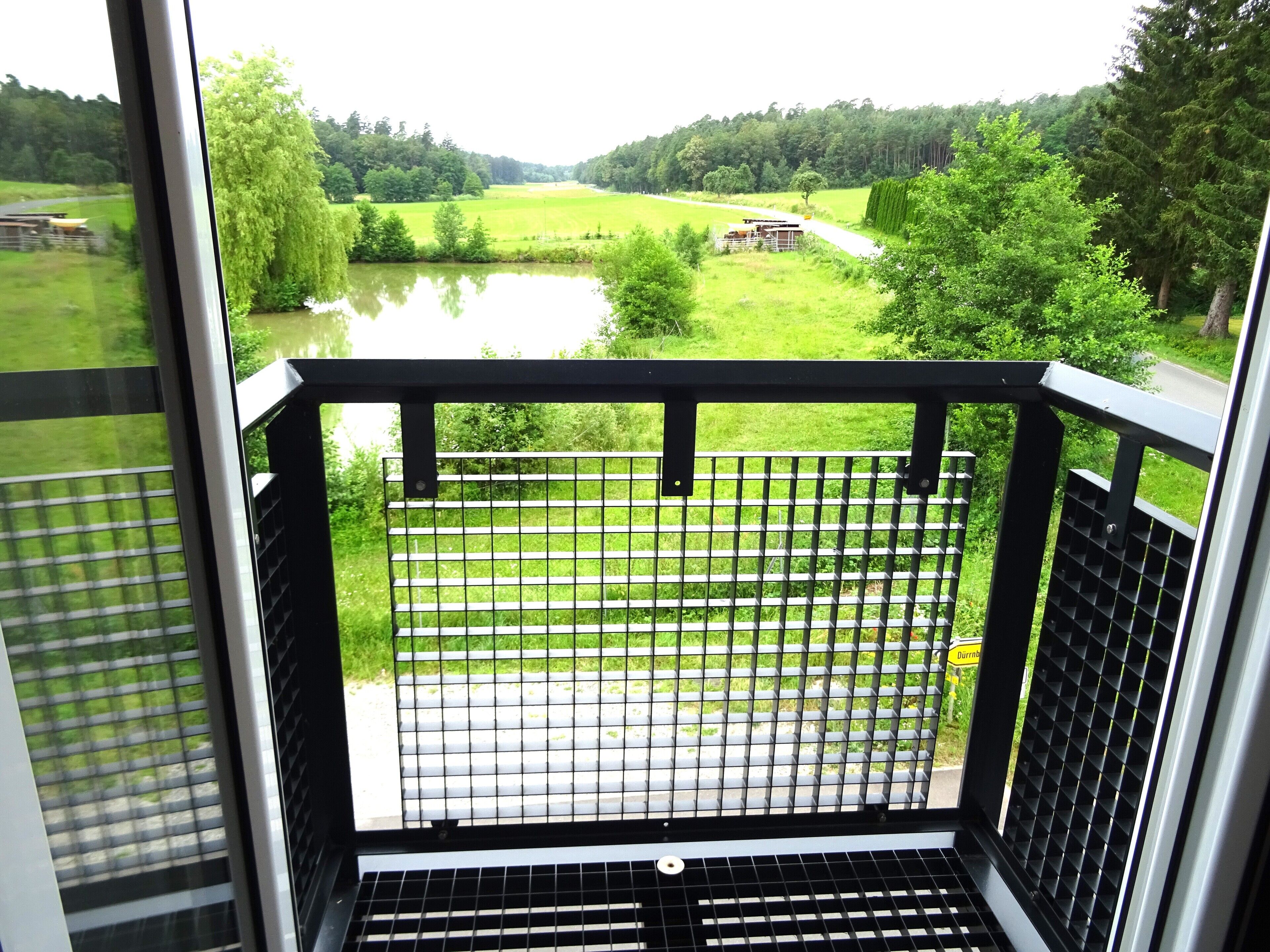 Uma varanda com grade de metal, vista para uma paisagem verde com um lago e árvores.