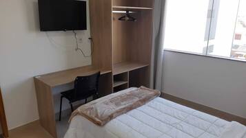 Quarto com cama, escrivaninha, cadeira, televisão na parede e janela.