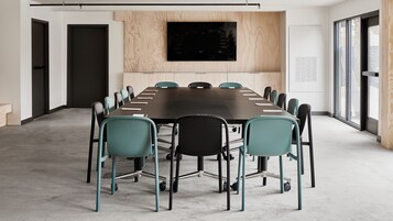 Uma sala de conferência com uma mesa comprida, cadeiras azul-esverdeadas, uma TV fixada na parede e grandes janelas.