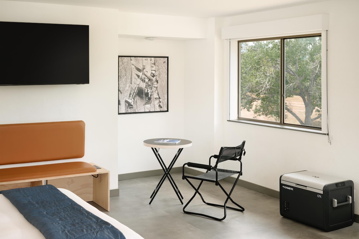 Quarto moderno e minimalista com cama, escrivaninha, cadeira, mesinha, quadro emoldurado e janela com vista para árvores.