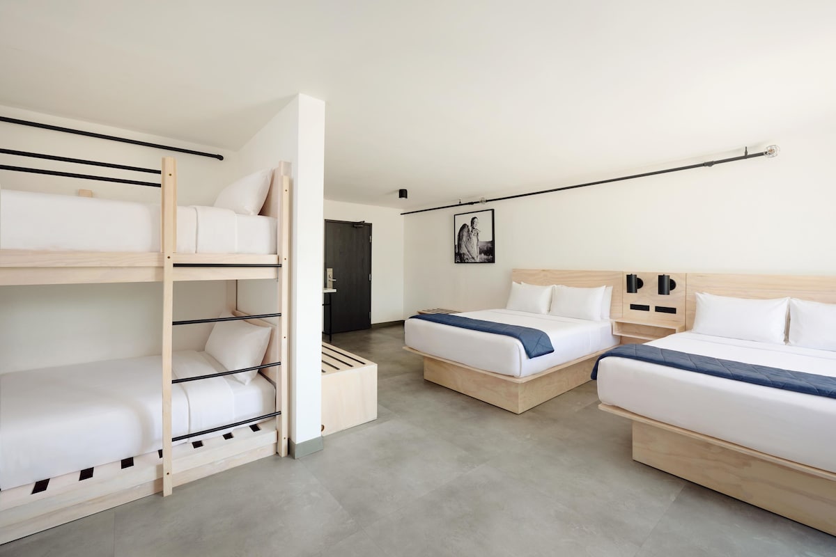 Quarto de hotel com beliches, uma cama de solteiro e cabeceira com compartimentos de armazenamento.