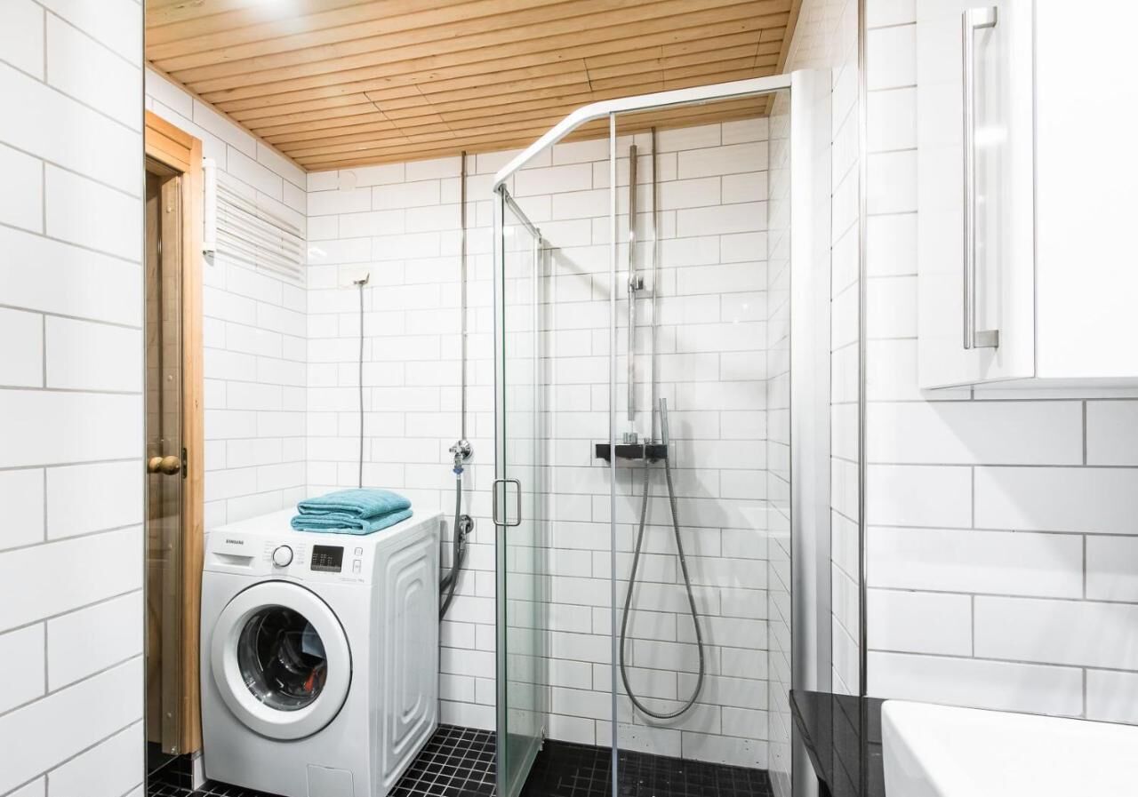Uma casa de banho compacta com máquina de lavar roupa, chuveiro em cabine de vidro e teto de madeira.