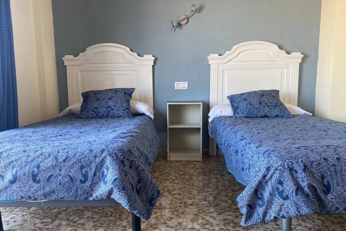 Quarto com duas camas, cada uma com cabeceira branca e roupa de cama estampada em azul. Há uma pequena mesa de cabeceira entre as camas.