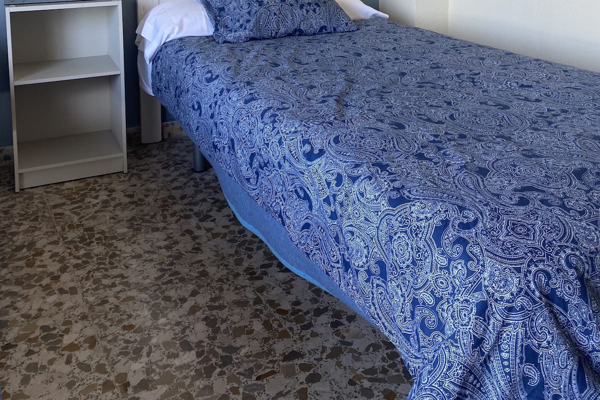 Uma cama de solteiro com cabeceira branca e edredom azul estampado. Um criado-mudo com abajur fica ao lado da cama.