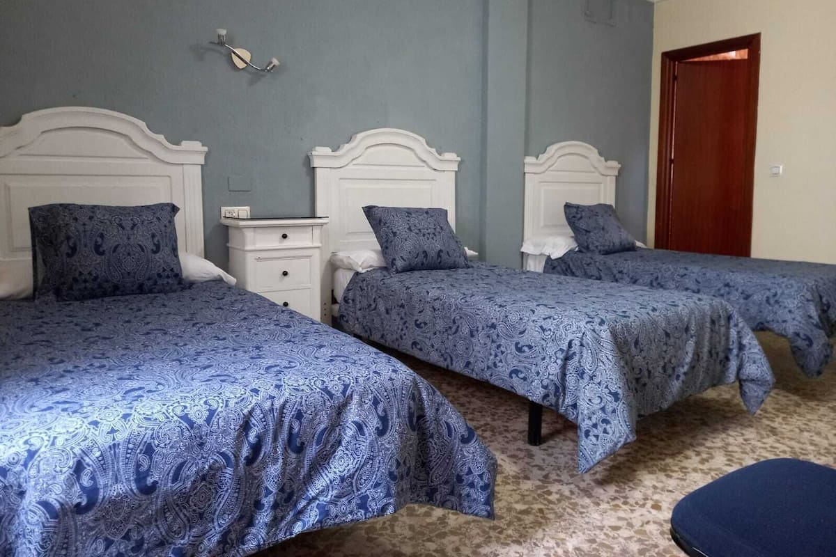 Quarto com três camas, cada uma com cabeceira branca e roupa de cama estampada em azul. Há uma porta de madeira e uma pequena mesa de cabeceira branca com uma luminária.