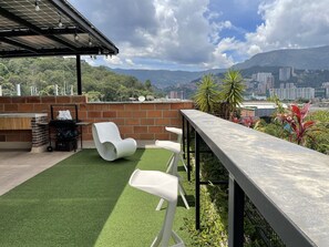 Terraço no telhado com grama artificial, cadeiras brancas, área de churrasco e vista para a cidade e montanhas.
