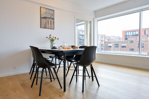 Uma área de refeições com mesa e cadeiras, um vaso com flores e vista para a cidade através de grandes janelas.