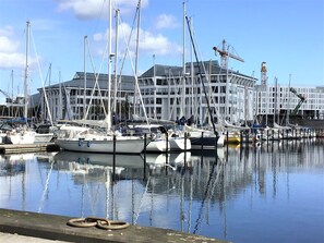 Uma marina com vários veleiros atracados, um edifício com arquitetura moderna ao fundo e águas calmas refletindo os barcos.