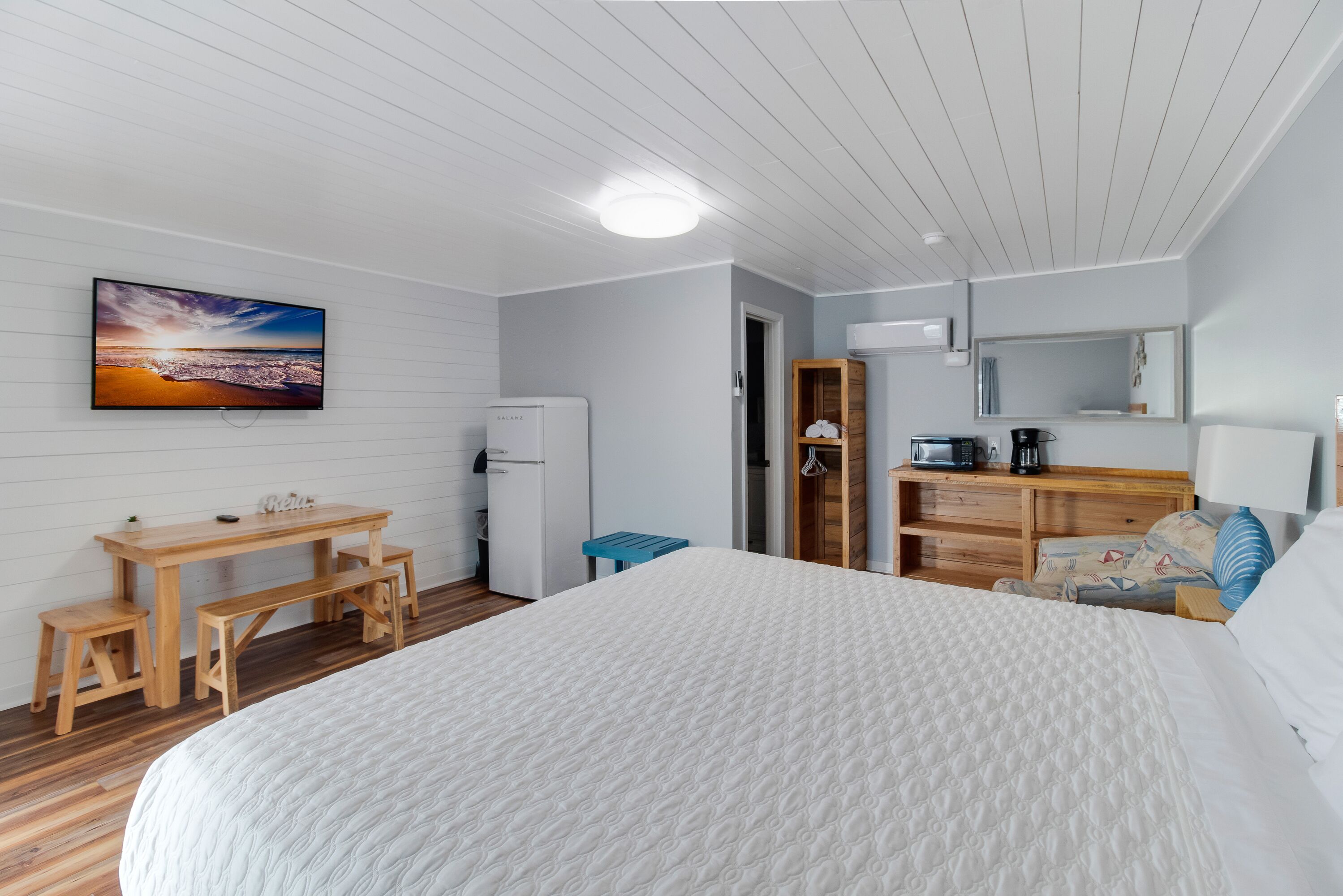Quarto com uma cama grande, uma escrivaninha de madeira com cadeira, uma mesinha e um banco. Há uma geladeira, micro-ondas e cafeteira na escrivaninha. Um espelho está pendurado na parede acima da cama.