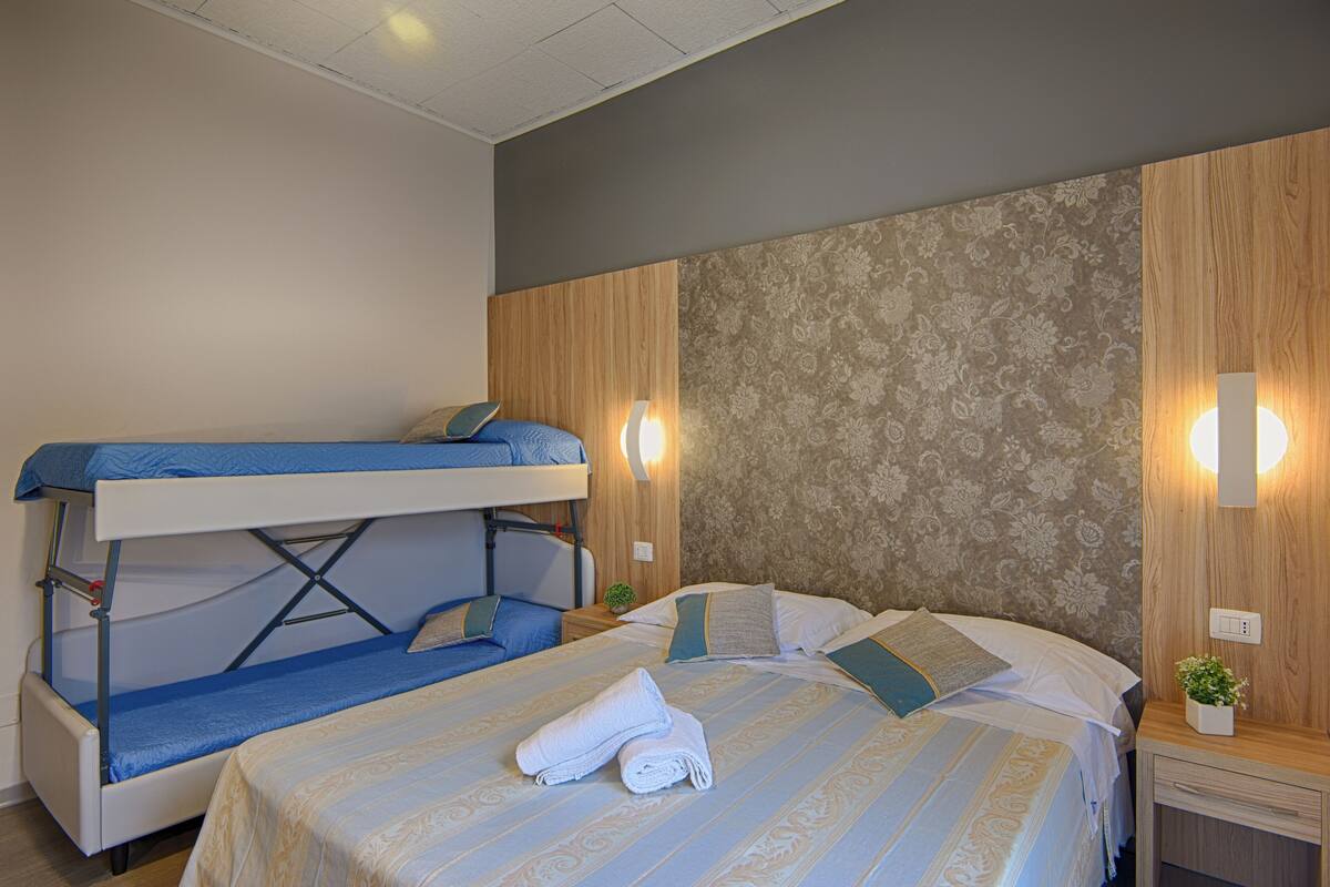 Quarto de hotel com uma cama de solteiro, uma cama individual com roupa de cama listrada, uma mesa de cabeceira com abajur e um luminário fixado na parede.