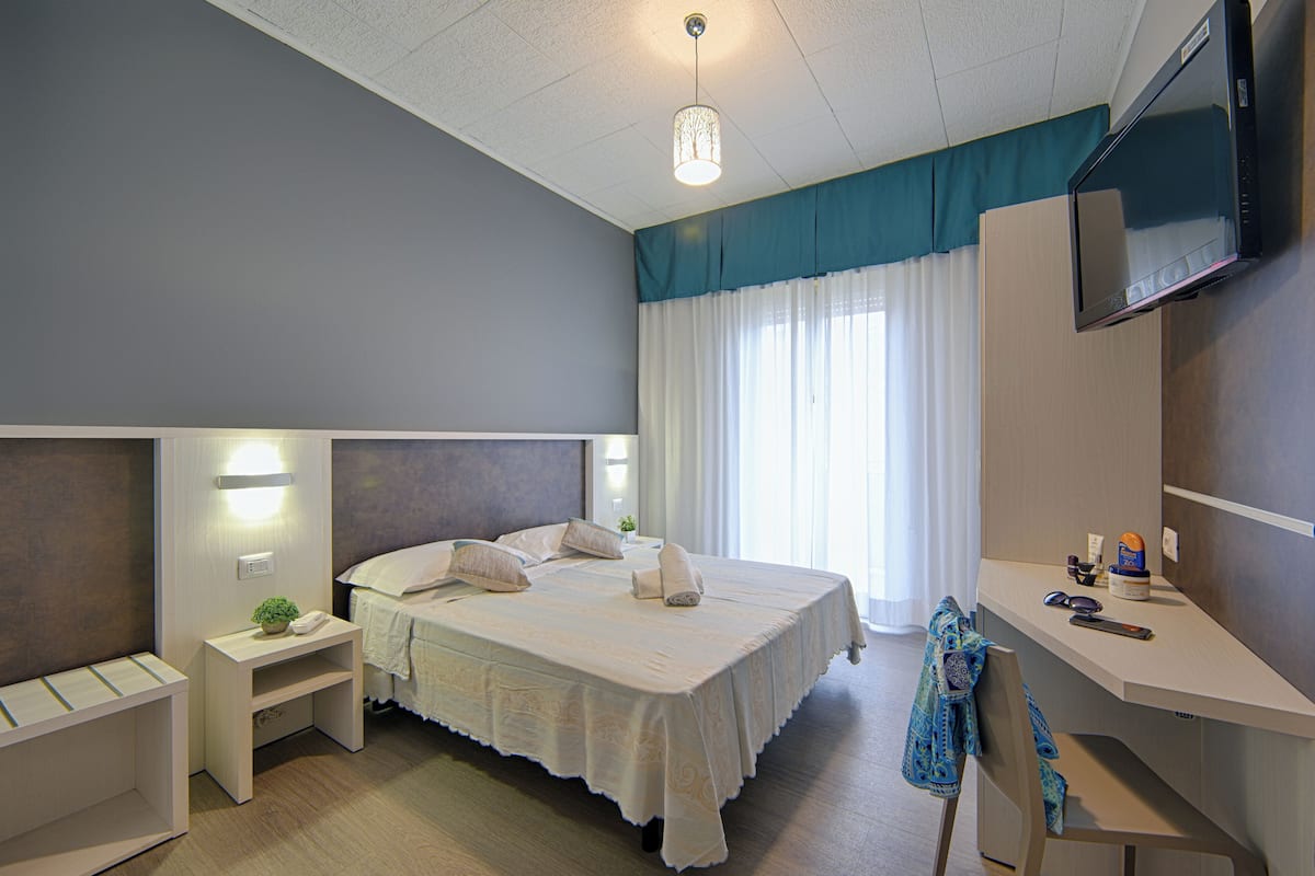 Quarto de hotel com cama, mesas de cabeceira, escrivaninha, televisão e janela com cortinas.