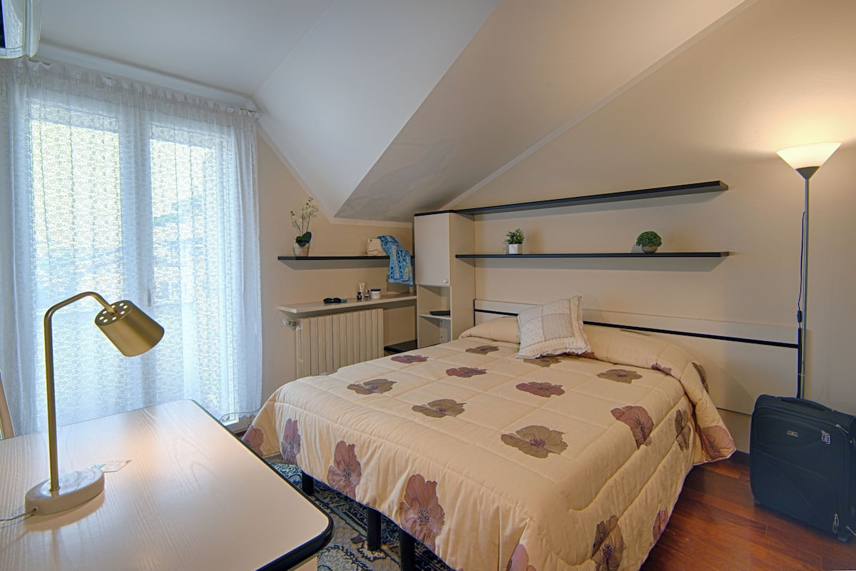 Quarto com cama, mesa de cabeceira, luminária e estantes com plantas.