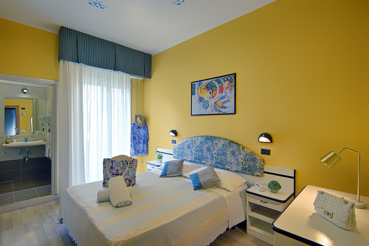 Quarto de hotel com cama, mesas de cabeceira, um espelho, janela com cortinas e um quadro na parede.