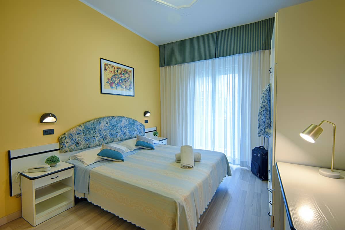 Quarto com cama, mesas de cabeceira, abajur, mala e um quadro na parede.