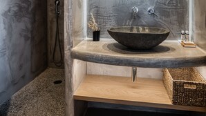 Uma casa de banho moderna com uma grande lavatório de pedra escura e arredondada, um móvel de banheira em madeira, chuveiro com porta de vidro e um espelho grande com moldura circular.