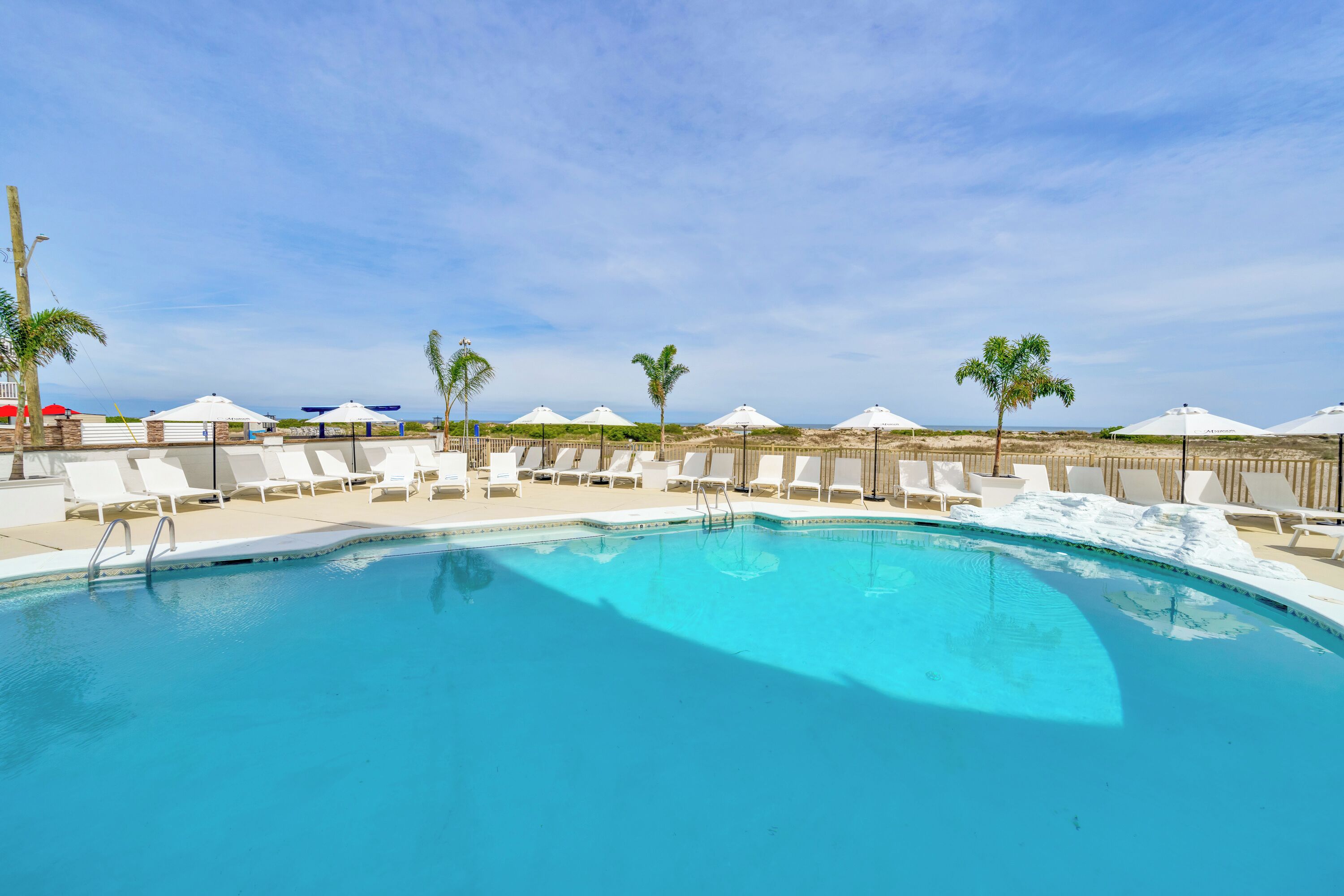 Piscina com espreguiçadeiras brancas e guarda-sóis, rodeada por palmeiras.