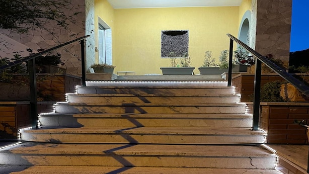 Uma escadaria com um padrão em zigue-zague de sombras, levando a um patamar com uma parede de pedra e uma pequena janela.
