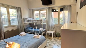 Quarto com beliche, escrivaninha, cadeira, televisão e janela com cortinas.