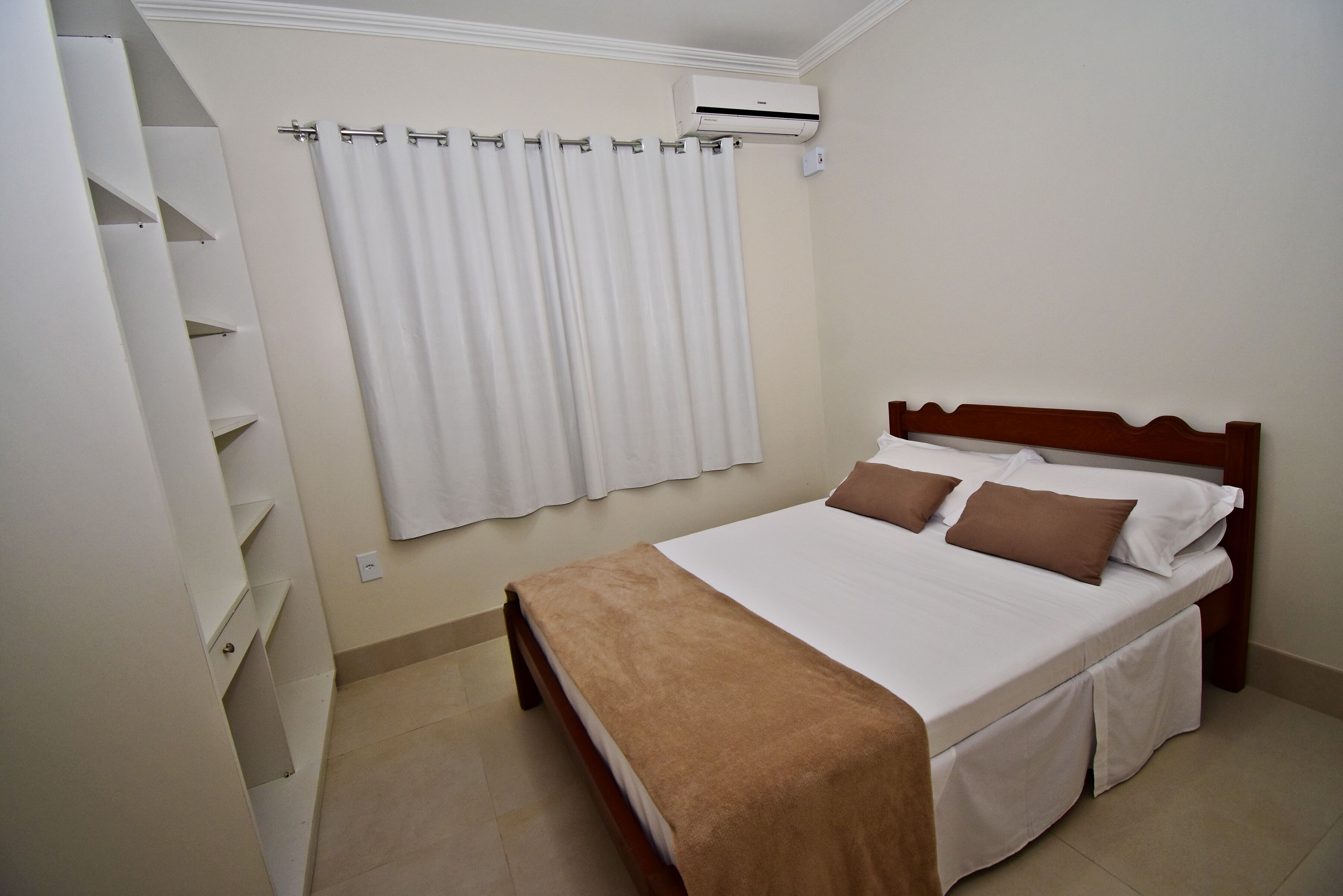 Quarto com cama, janela com cortinas, ar-condicionado fixado na parede e armário embutido.