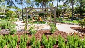 Um parque com uma pérgula de madeira, um parquinho e áreas ajardinadas com arbustos e árvores.