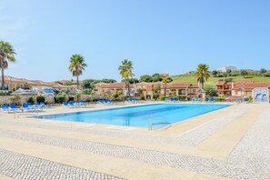 Piscina com espreguiçadeiras e palmeiras, cercada por edifícios com telhados de terracota.