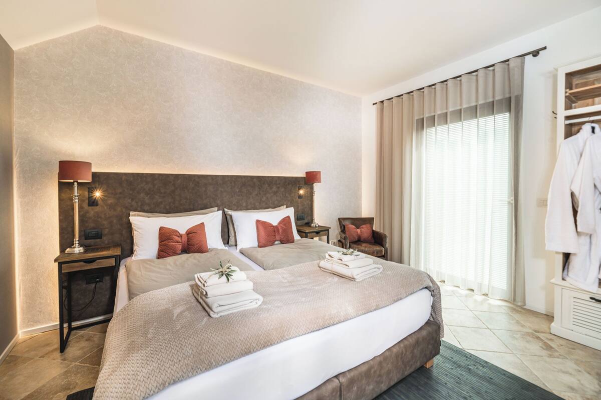 Quarto de hotel com uma cama grande, duas mesas de cabeceira, uma cadeira, um guarda-roupa e uma janela com cortinas.