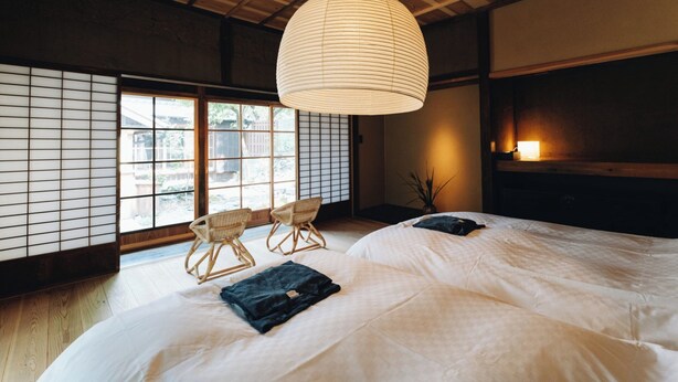 Um quarto tradicional japonês, com piso de tatame, uma cama grande com roupa de cama branca, duas cadeiras de palha, um lanterna de papel pendente e vista através de um painel shoji.
