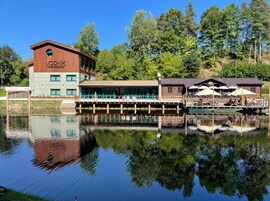 Um edifício chamado VODNIK com fachada de madeira, um cais que se estende por um lago e uma área de restaurante com guarda-sóis.