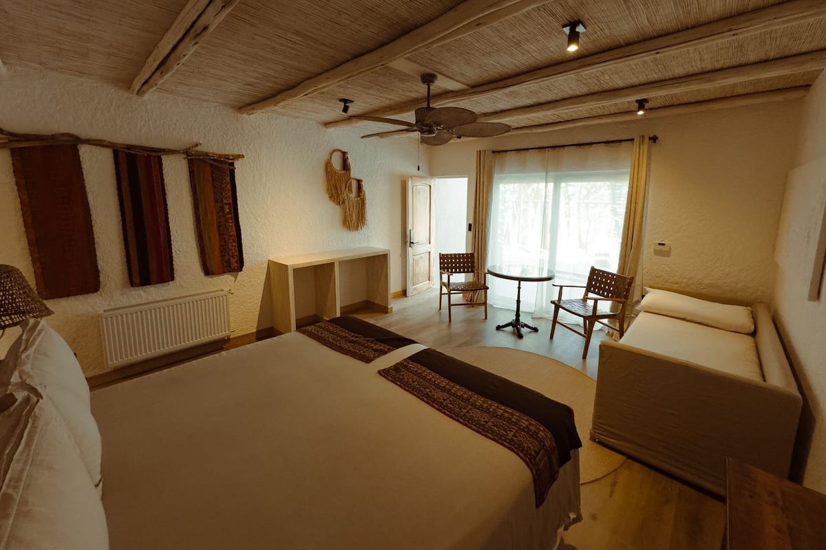 Um quarto com uma cama grande, uma escrivaninha, duas cadeiras, um ventilador de teto e uma janela com cortinas.