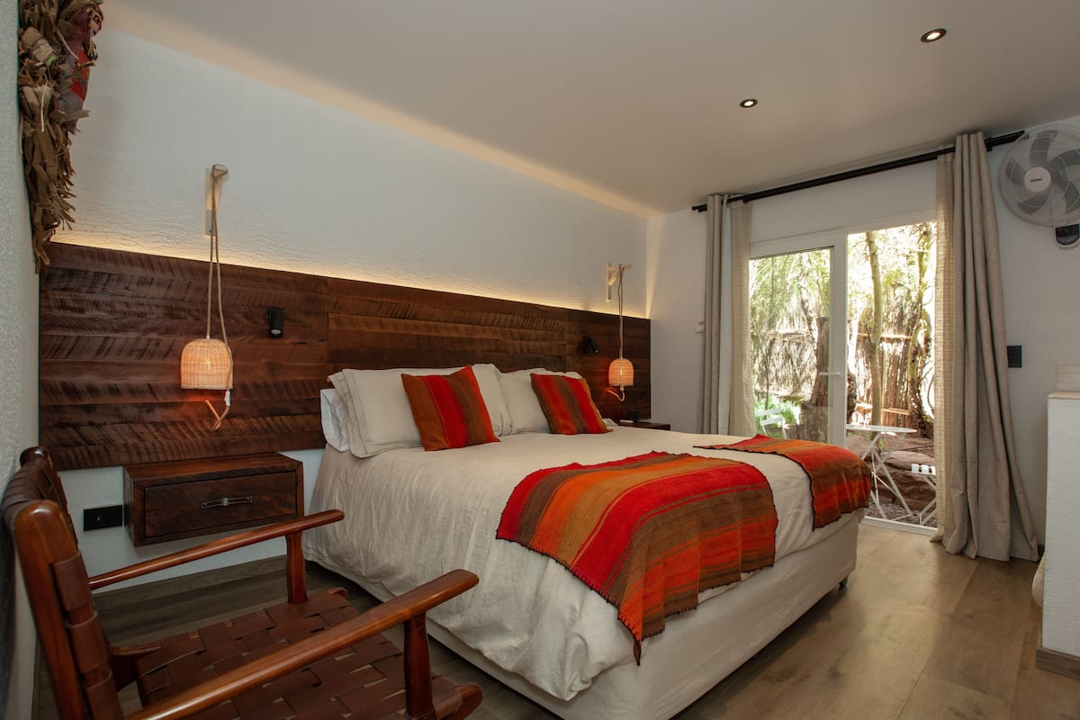 Quarto com uma cama grande, cabeceira de madeira, mesas de cabeceira, uma cadeira e vista para o exterior por uma porta deslizante.