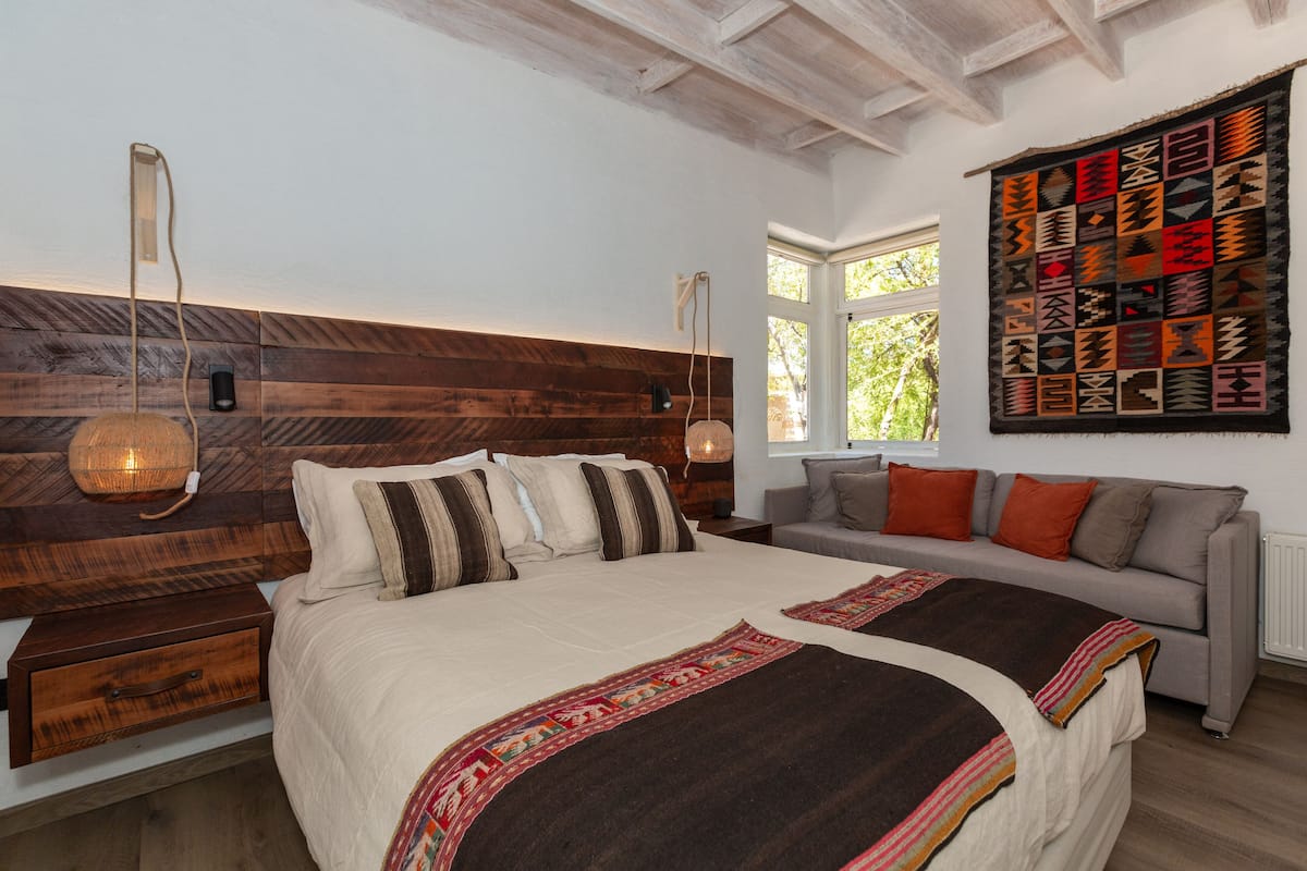 Quarto com cabeceira de madeira, cama grande com várias almofadas, sofá cinza e um pendurar colorido na parede.