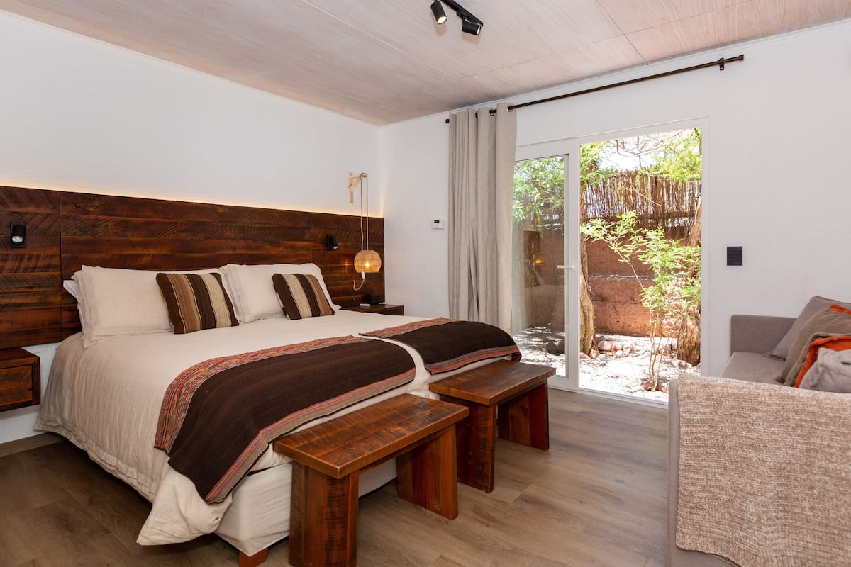 Quarto com uma cama grande, cabeceira de madeira, mesas de cabeceira, um banco e vista de um jardim por uma porta de vidro deslizante.