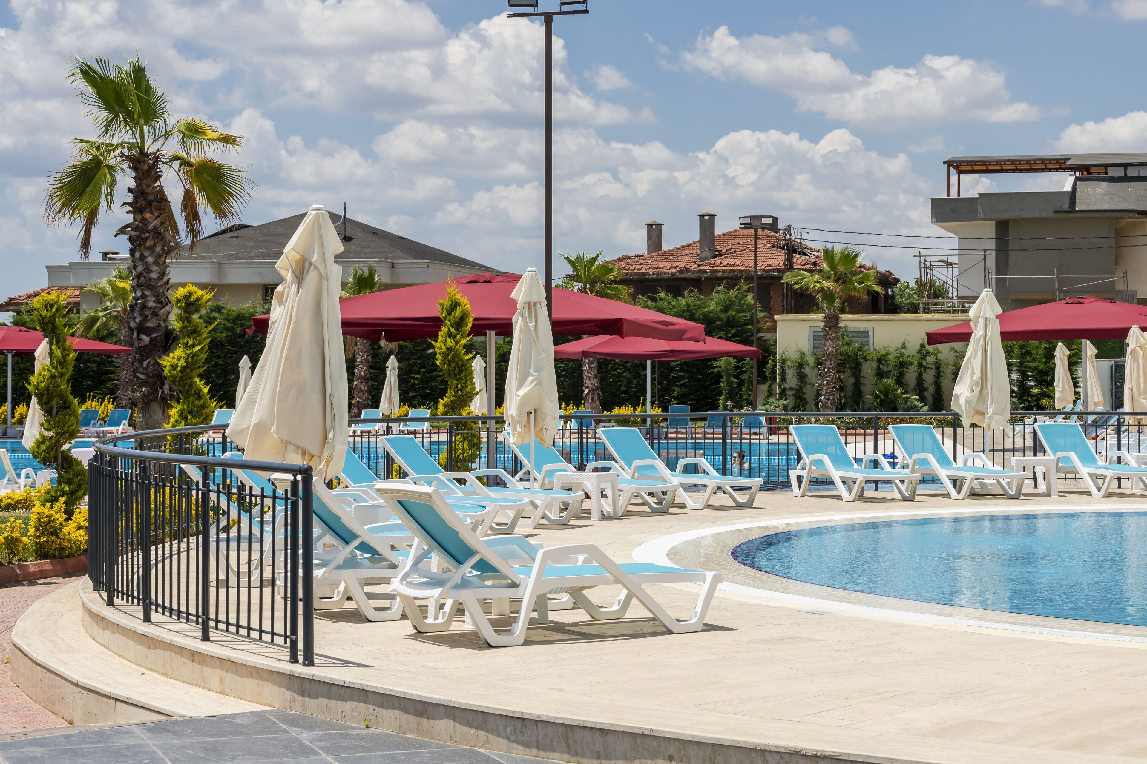 Área de piscina com espreguiçadeiras azuis e guarda-sóis vermelhos, cercada por palmeiras e um edifício ao fundo.