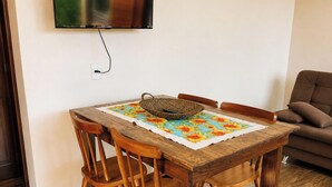 Uma área de refeições com uma mesa de madeira, cadeiras, uma cesta de palha e um jogo de mesa floral. Uma televisão de tela plana está fixada na parede acima da mesa.