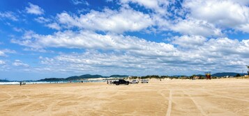 Uma extensa praia de areia com céu claro e montanhas ao longe.