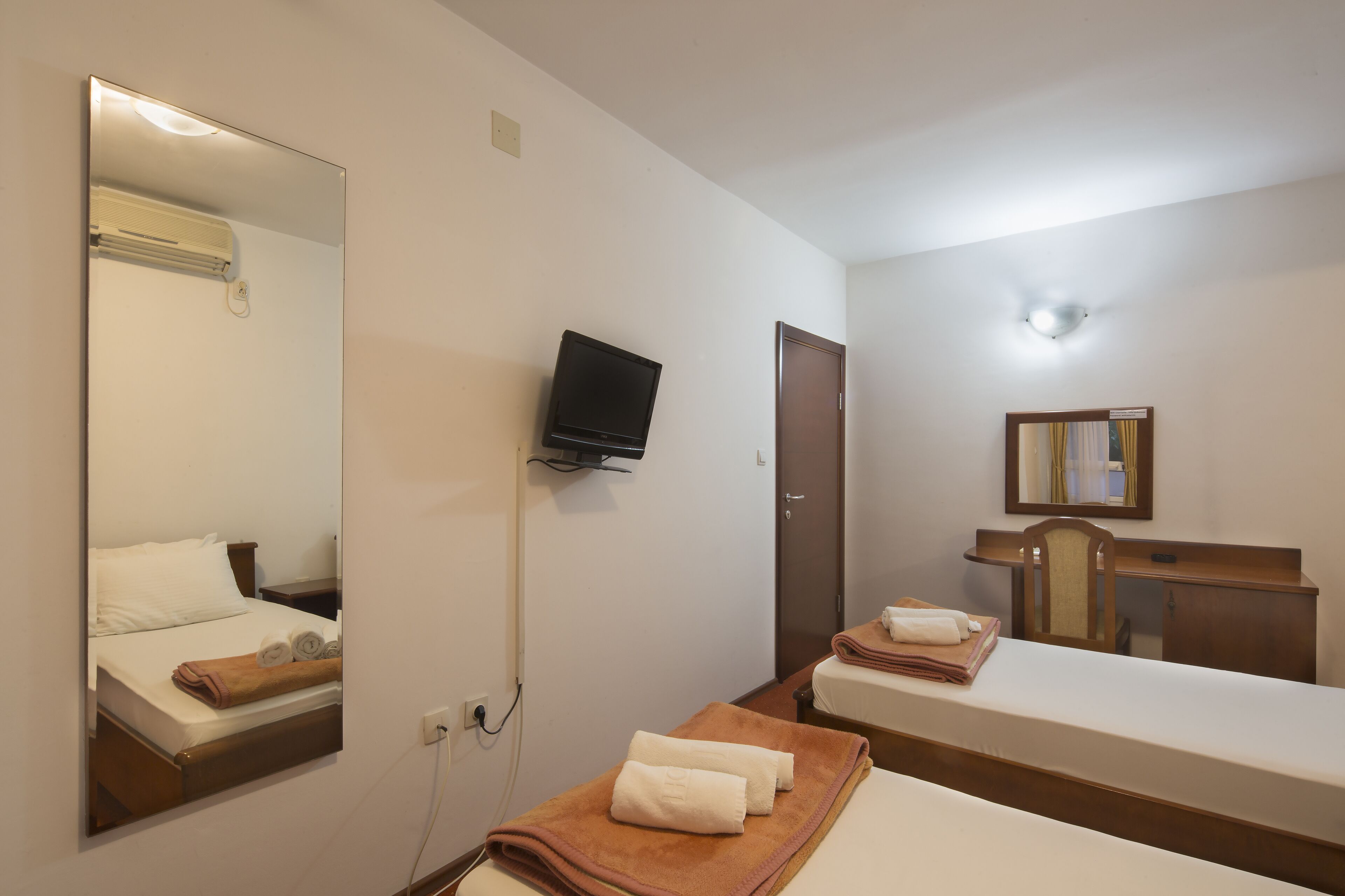 Quarto de hotel com cama, escrivaninha, televisão, espelho e porta do banheiro.