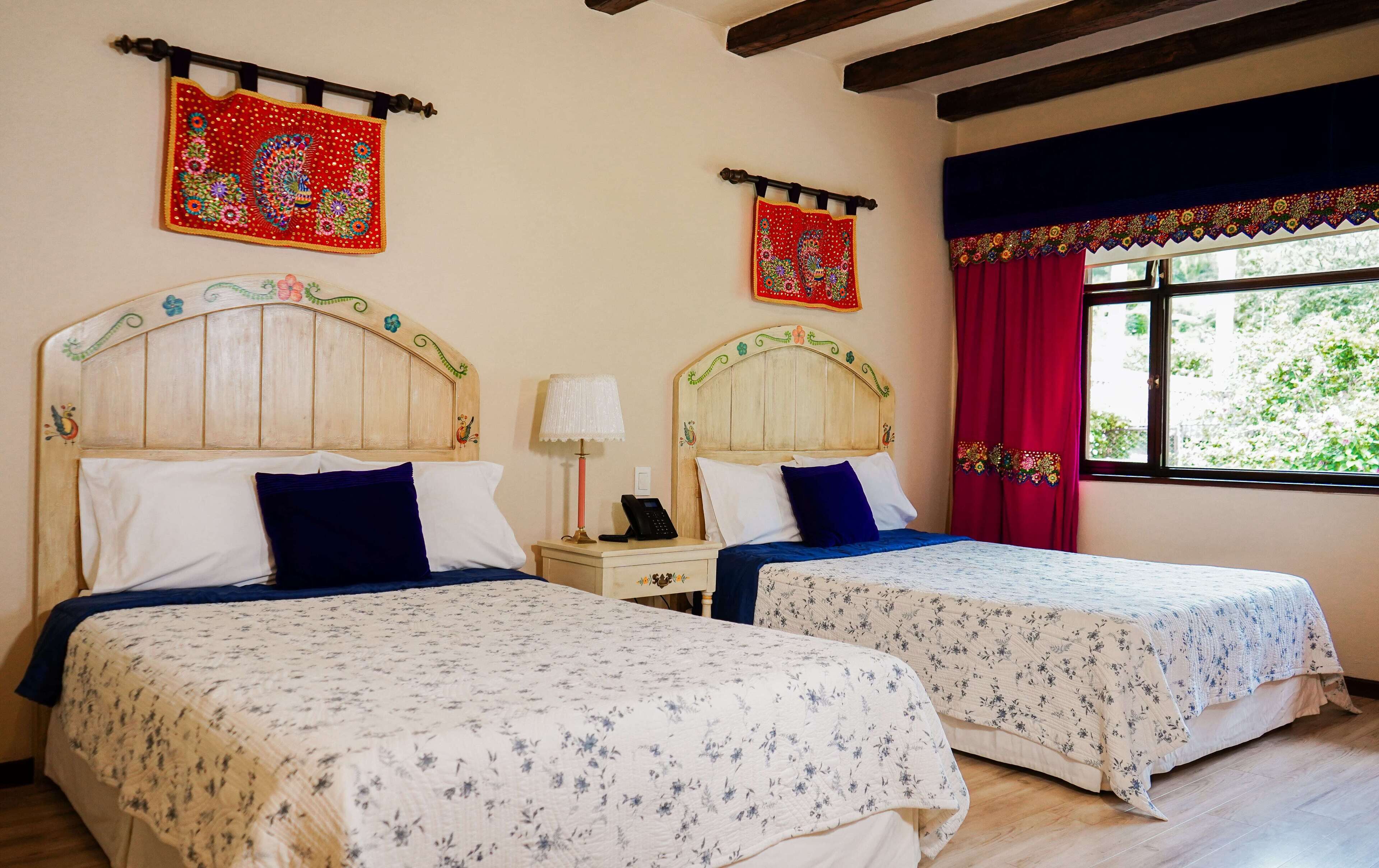 Quarto com duas camas, cada uma com colcha floral e almofadas combinando, luminária de cabeceira, telefone na mesinha de cabeceira e janela com cortinas vermelhas.