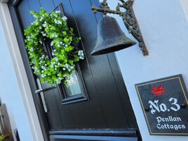 Uma porta preta com uma grinalda de flores, um sino e uma placa com a inscrição 'No. 3 Penllan Cottages'.